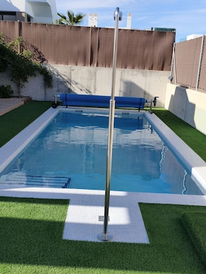 Piscina retangular com um poste de metal no centro, cercada por grama sintética e um deck de concreto.