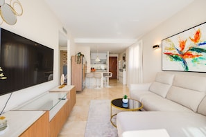 Uma sala de estar moderna com televisão de tela plana, um sofá, uma mesa de centro e uma área de jantar ao fundo.
