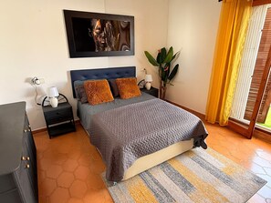 Quarto com cama, mesas de cabeceira, televisão na parede, planta em vaso e janela com cortinas.