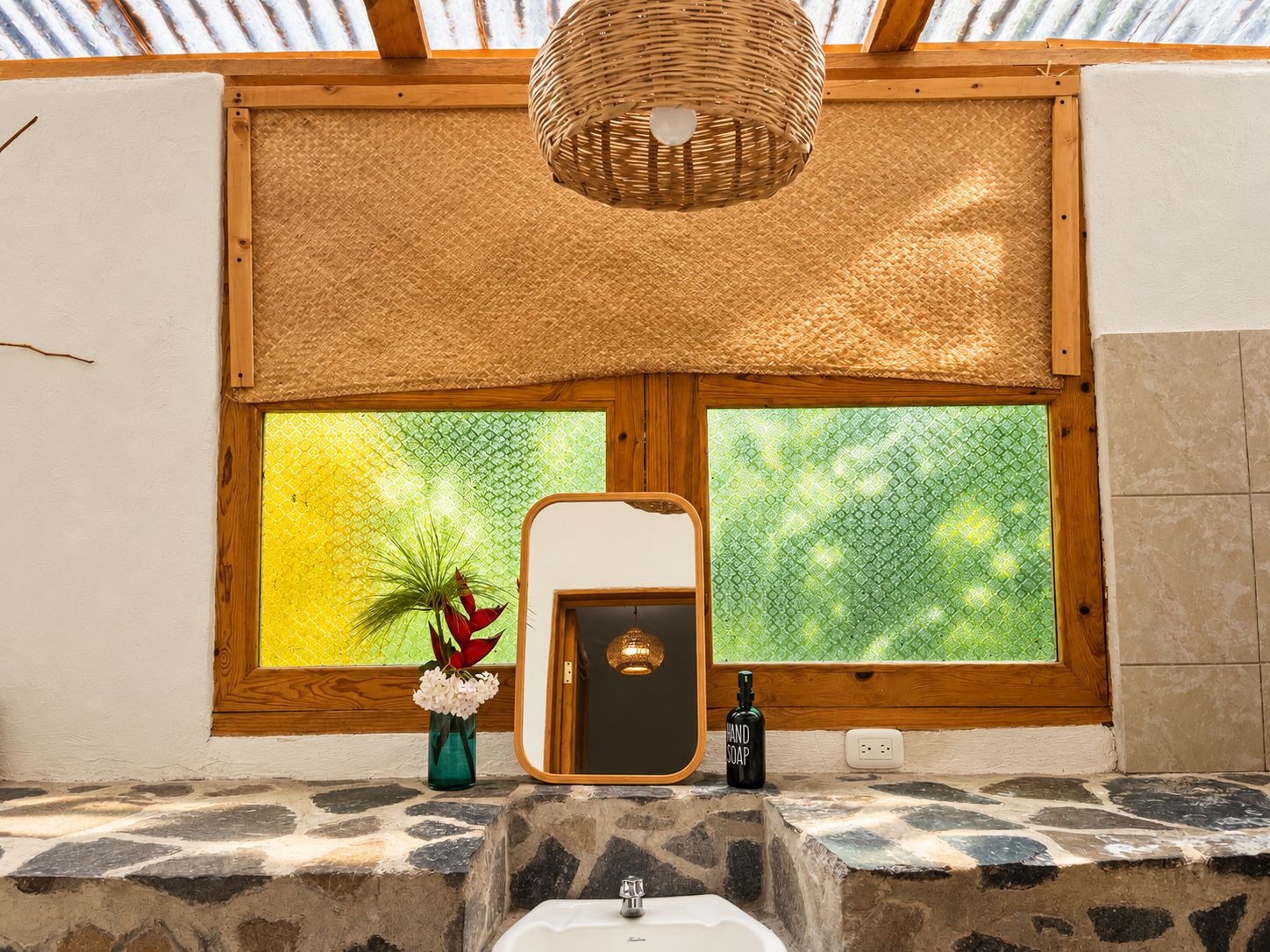 Casa de banho com lavatório de pedra, espelho, dispensador de sabonete líquido e janela com vista para vegetação.