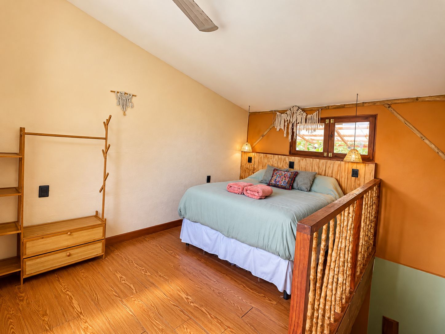 Quarto com cama, escada de madeira, janela com decoração em macramé e corrimão de madeira.