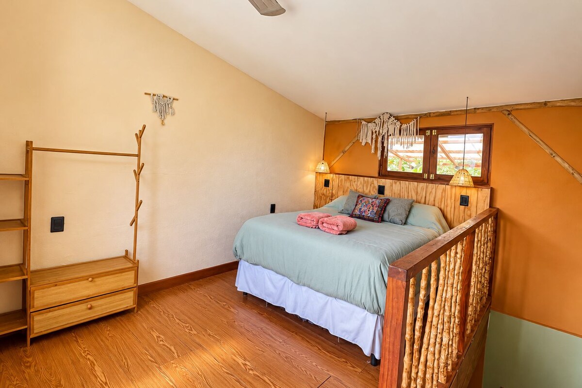 Quarto com cama, escada de madeira, janela com decoração em macram é e corrimão de madeira.