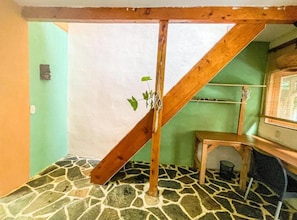 Um quarto com teto inclinado, estrutura de vigas de madeira, piso de pedra, uma mesa pequena e uma cadeira.