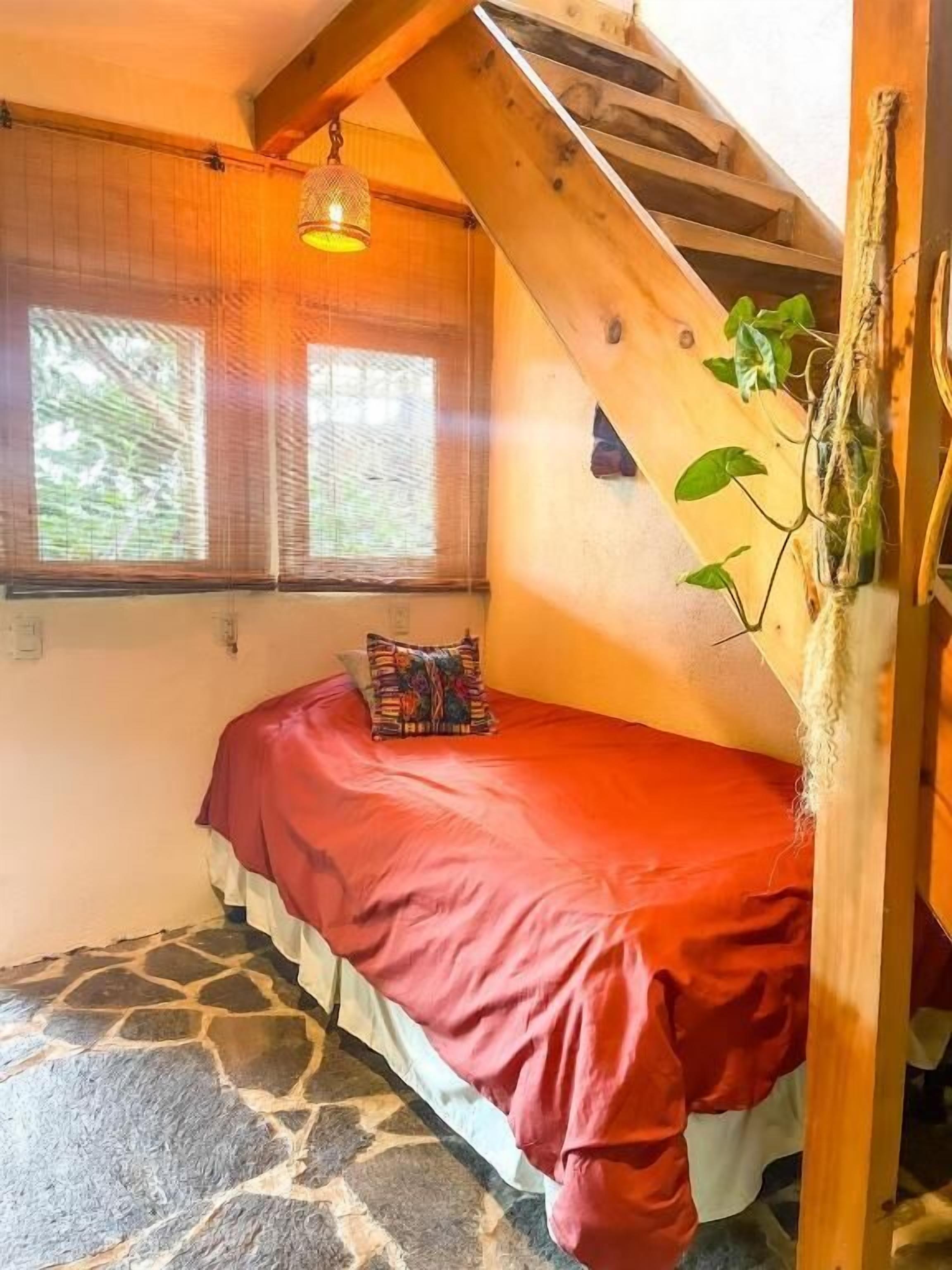 Uma cama de solteiro com colcha vermelha em um quarto com teto inclinado e vigas de madeira.