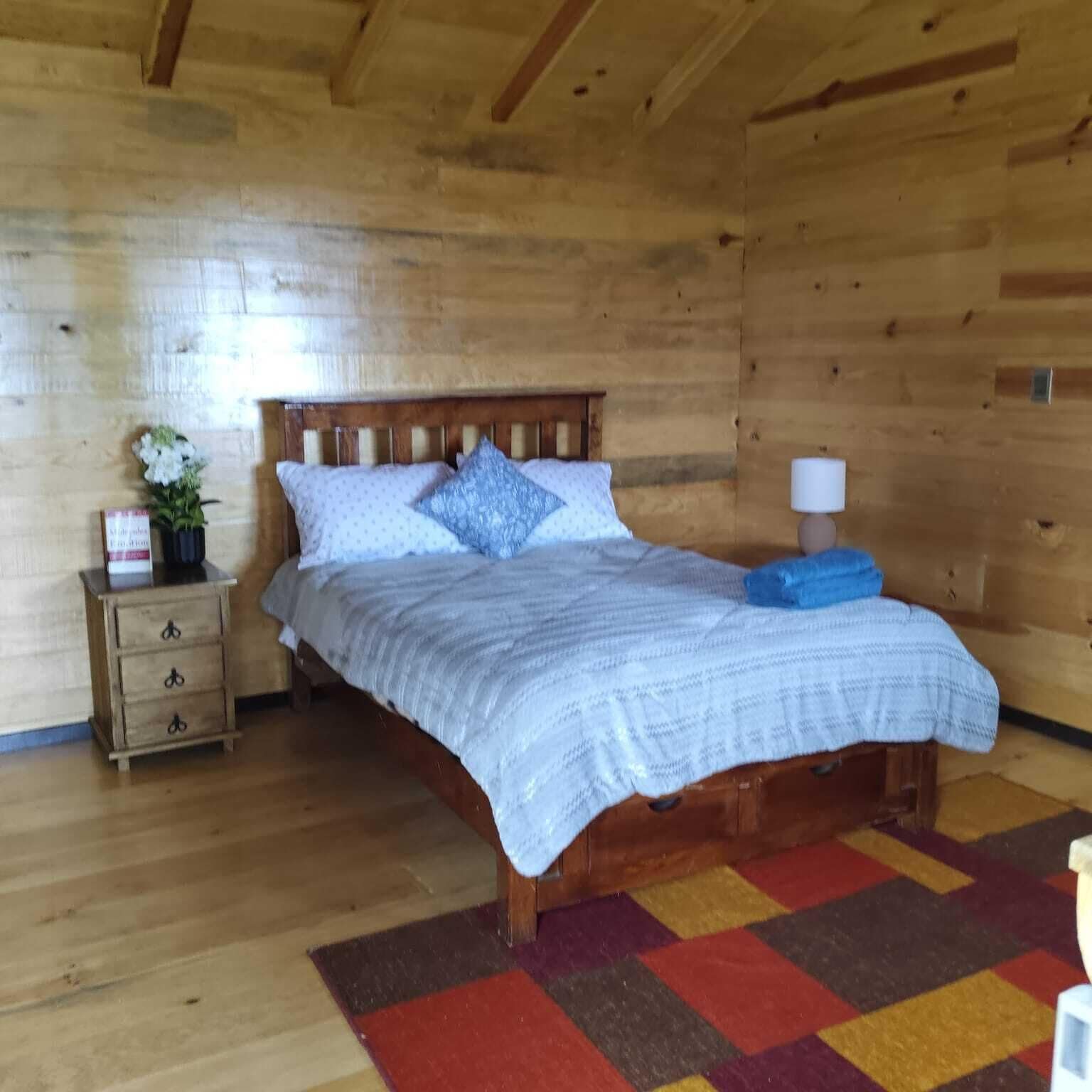 Quarto de cabana de madeira com cama, mesa de cabeceira, abajur e tapete colorido.