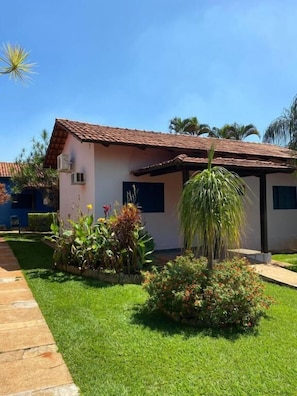 Um edifício térreo com telhado de cerâmica, um pequeno jardim com diversas plantas e um caminho pavimentado que leva à entrada.