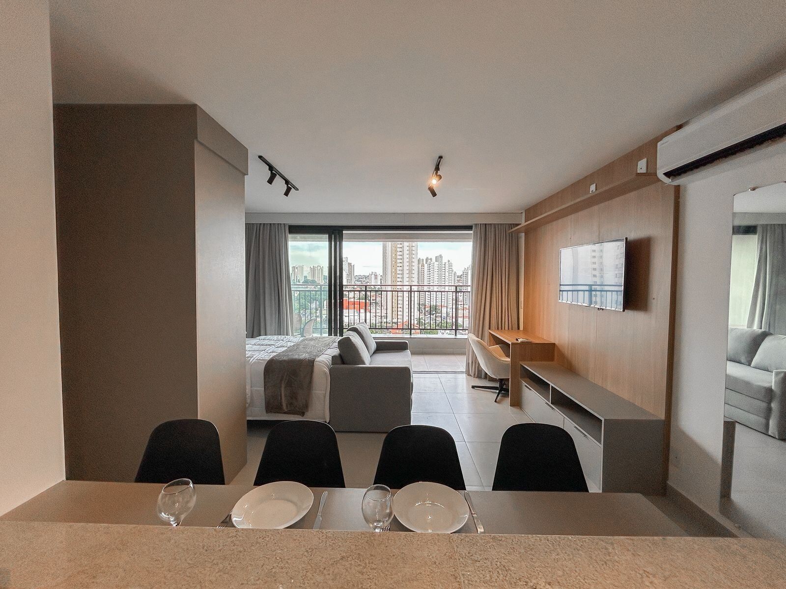 Quarto de hotel moderno com área de jantar, sofá, TV e varanda com vista para a cidade.