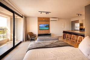 Quarto de hotel moderno com uma cama grande, televisão, secretária com banquetas altas e varanda com cortinas.