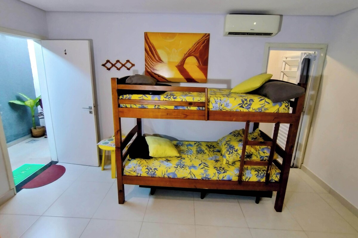 Uma cama beliche com roupa de cama florida, um banquinho pequeno, uma planta em vaso e uma porta que dá para outro quarto.