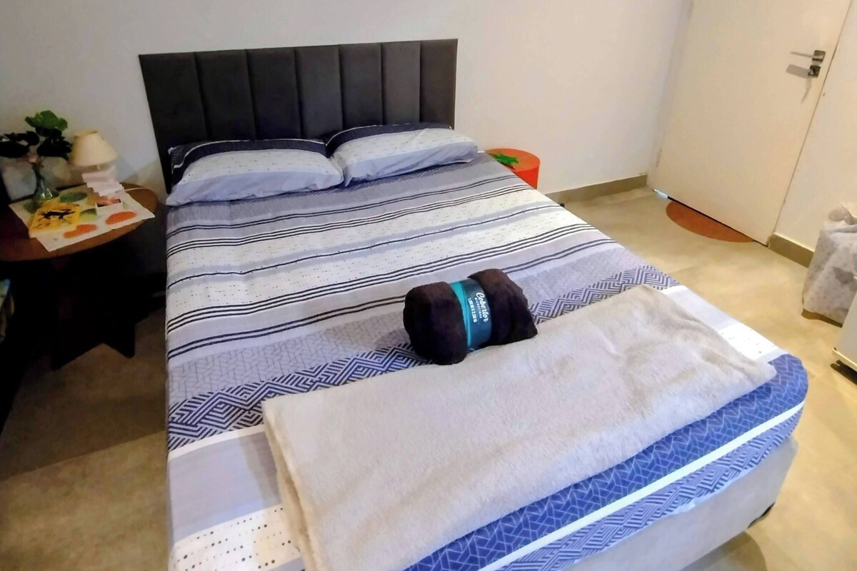 Cama bem arrumada com colcha estampada, toalha dobrada e uma caminha para cachorro na cama. Três quadros emoldurados na parede acima da cama.