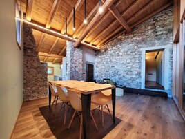 Interior rústico com mesa e cadeiras de madeira, paredes de pedra e vigas de madeira aparentes.