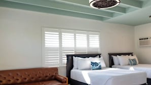 A hotel room with a brown leather sofa, two beds with white bedding, a ceiling fan, and a window with blinds.