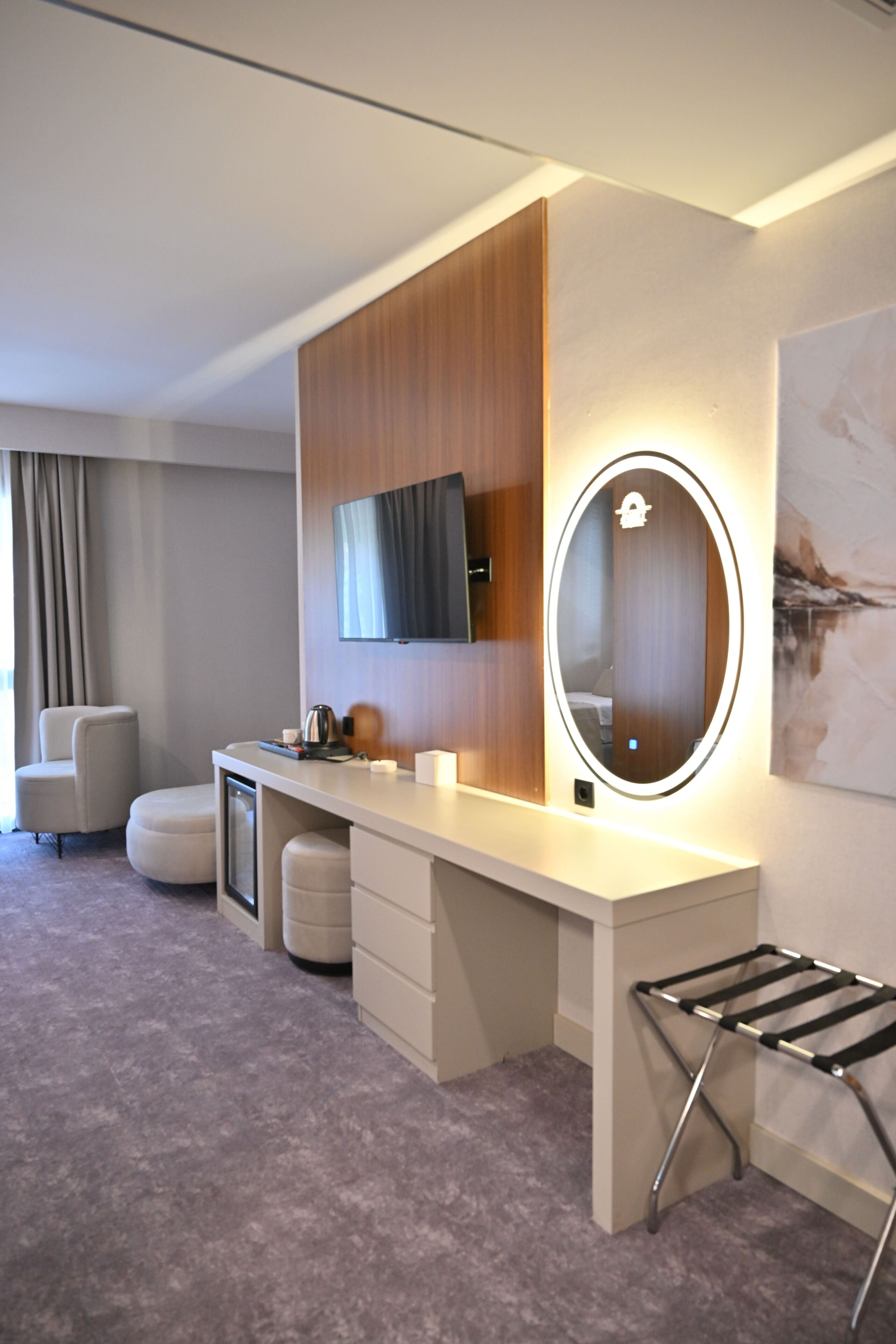 Quarto de hotel moderno com um espelho grande, TV de tela plana, escrivaninha com chaleira e área de estar com poltrona e apoio para os pés.