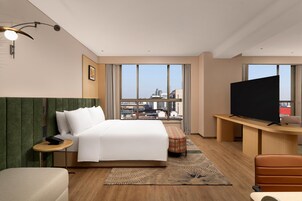 Quarto de hotel com uma cama, uma escrivaninha com televisão, uma cadeira, uma mesinha e vista para a paisagem urbana através de grandes janelas.