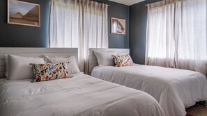 Quarto de hotel com duas camas, roupa de cama branca e vista para o deserto através de cortinas translúcidas.