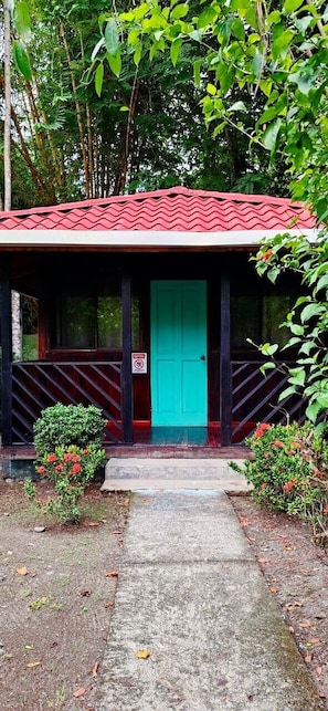 Um pequeno edifício com telhado vermelho, porta azul-turquesa e um caminho que leva até ele.