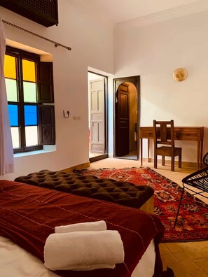 Quarto com cama, escrivaninha e cadeira. Há uma janela com persianas coloridas e uma porta que dá para outro quarto.