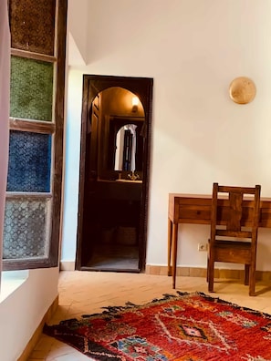 Quarto com escrivaninha de madeira, uma cadeira, tapete estampado e porta que dá para o interior escuro.