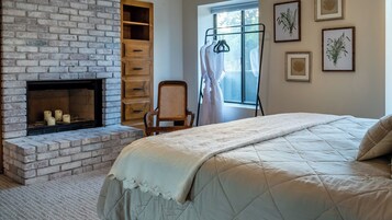 Um quarto com lareira de tijolos, uma cama com colcha acolchoada, uma cadeira de madeira e gravuras botânicas emolduradas na parede.