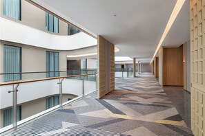 Corredor de hotel com carpete estampado, guarda-corpos de vidro e porta de madeira.