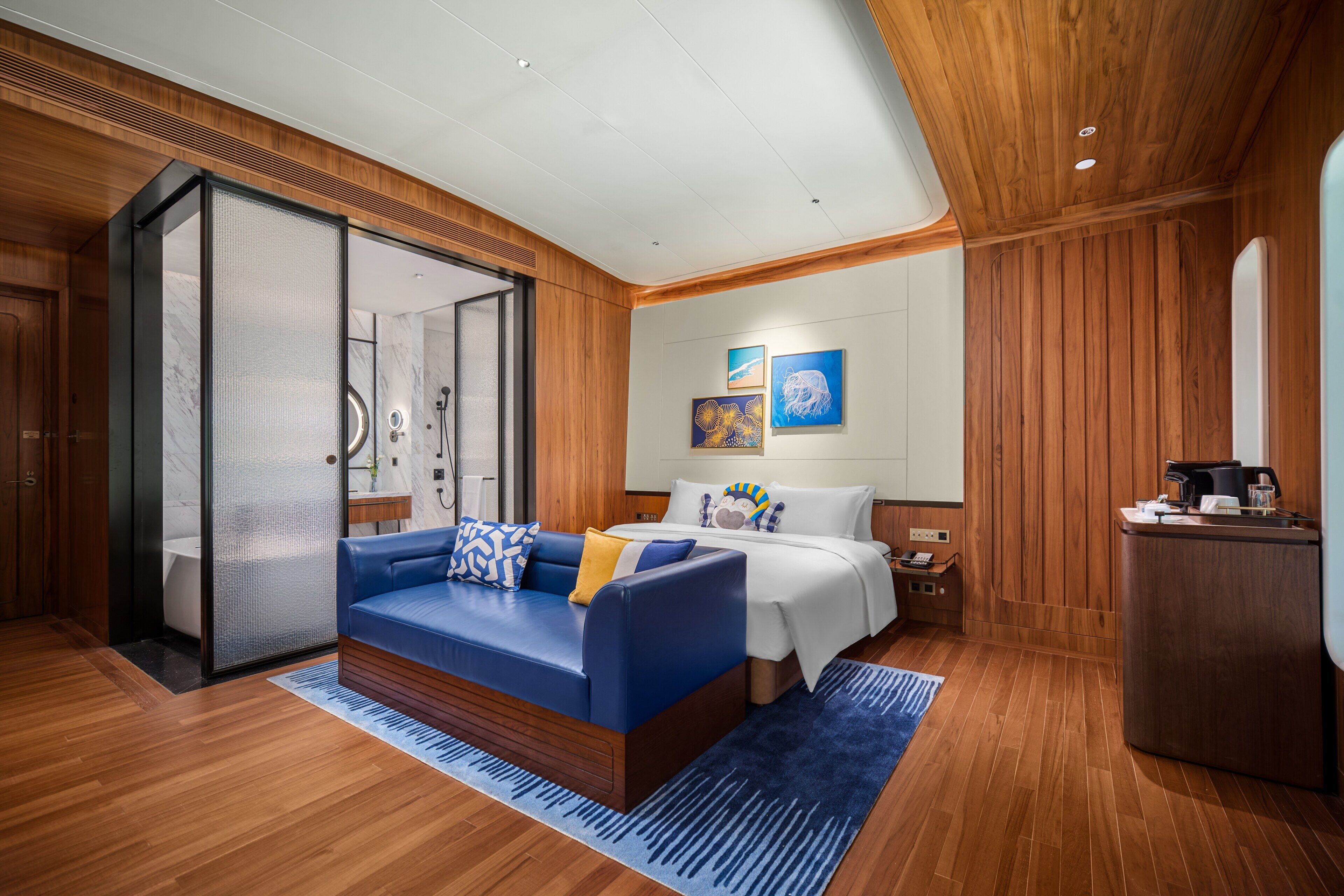 Quarto de hotel moderno com piso de madeira, um sofá azul, uma cama com roupa de cama branca e uma pequena escrivaninha com cafeteira.