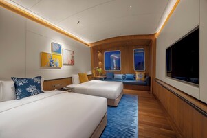 Quarto de hotel moderno com duas camas, obras de arte nas paredes, área de estar com vista e uma televisão grande.