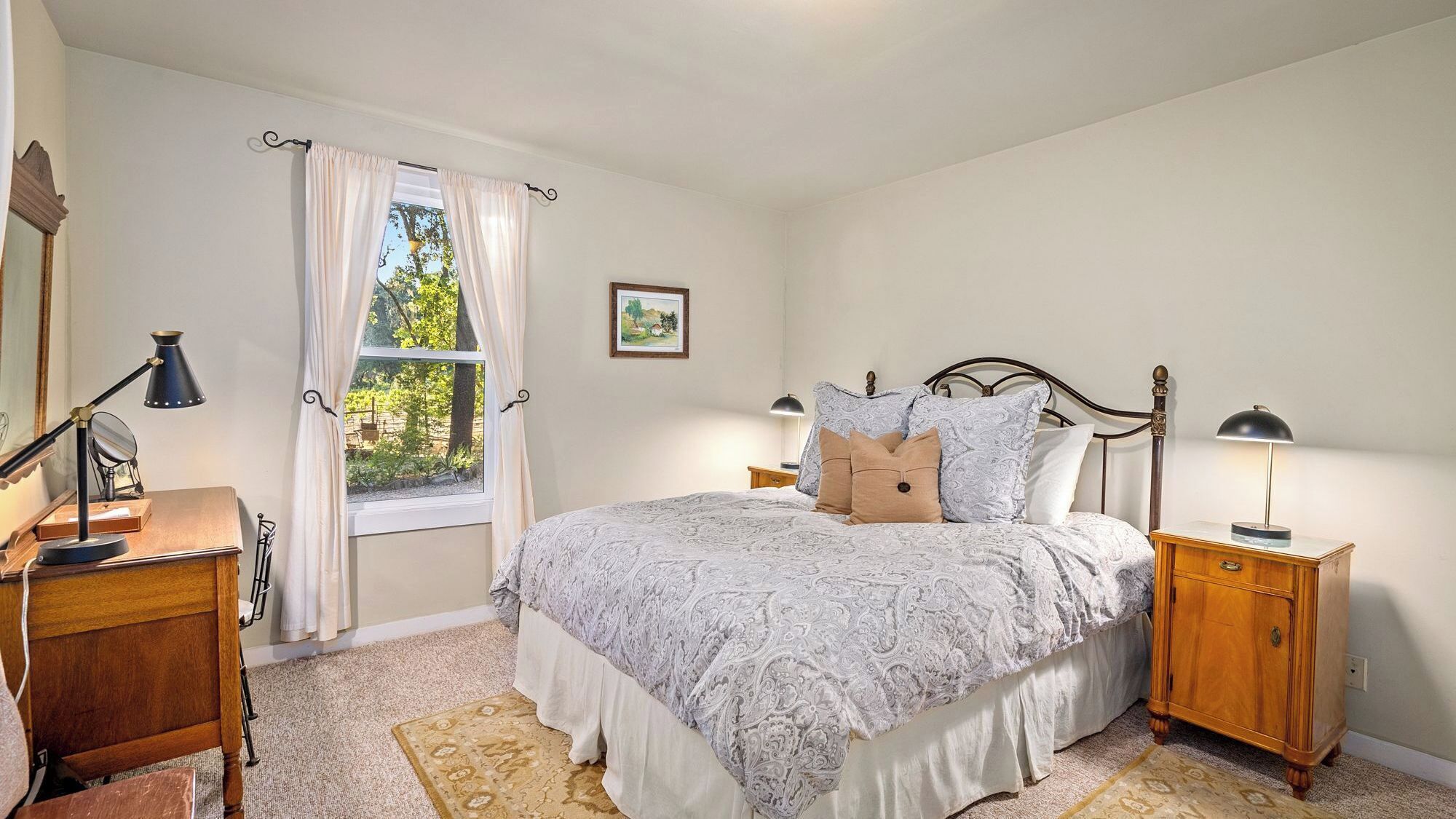 Quarto com cama, duas mesas de cabeceira, escrivaninha, cadeira, janela com cortinas e quadro na parede.
