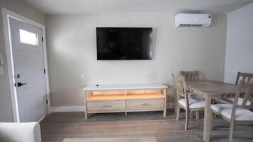 Um quarto com uma televisão branca fixada na parede, uma mesa de jantar de madeira com cadeiras, um banco branco com gavetas e um tapete estampado no chão.