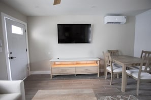Um quarto com uma televisão branca fixada na parede, uma mesa de jantar de madeira com cadeiras, um banco branco com gavetas e um tapete estampado no chão.