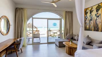Quarto com cama, escrivaninha de madeira, sofá e um quadro. Tem vista para a praia através de uma porta de vidro.
