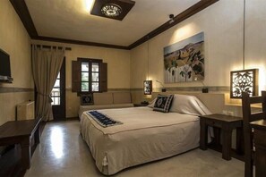 Quarto com cama, mesas de cabeceira, escrivaninha, cadeira, janela com cortinas e um quadro na parede.