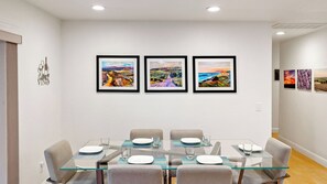 Uma sala de jantar com mesa de vidro, seis cadeiras e três quadros na parede.