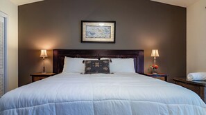 Quarto de hotel com uma cama grande, duas luminárias de cabeceira, uma obra de arte emoldurada na parede e uma janela com persianas.