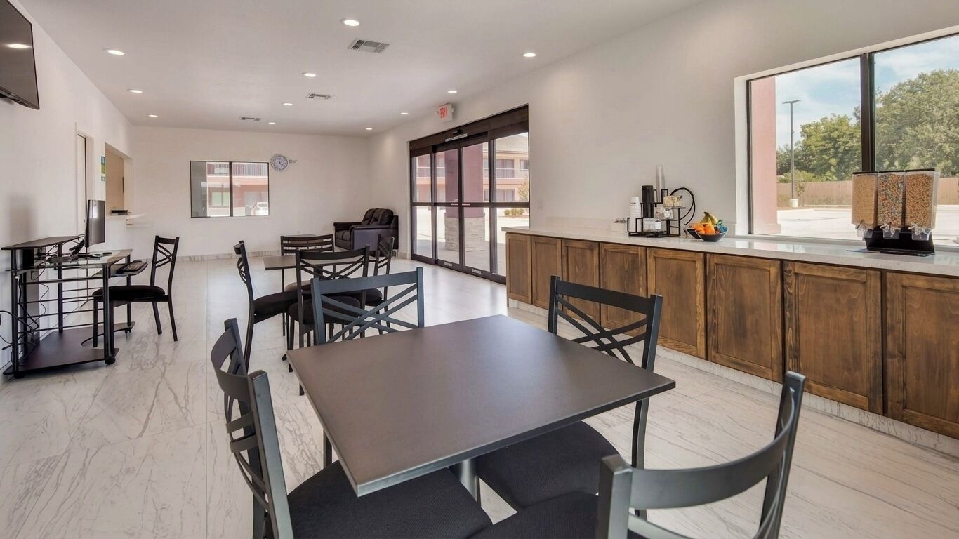 Uma área de jantar moderna com uma mesa comprida e cadeiras, um balcão de cozinha com equipamento para café e janelas amplas com vista para o exterior.