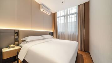 Quarto de hotel com uma cama grande, mesa de cabeceira com abajur, ar condicionado e janela com cortinas.