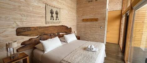 Quarto com cabeceira de madeira, cama com roupa de cama branca, mesa de cabeceira pequena com abajur e uma janela grande.