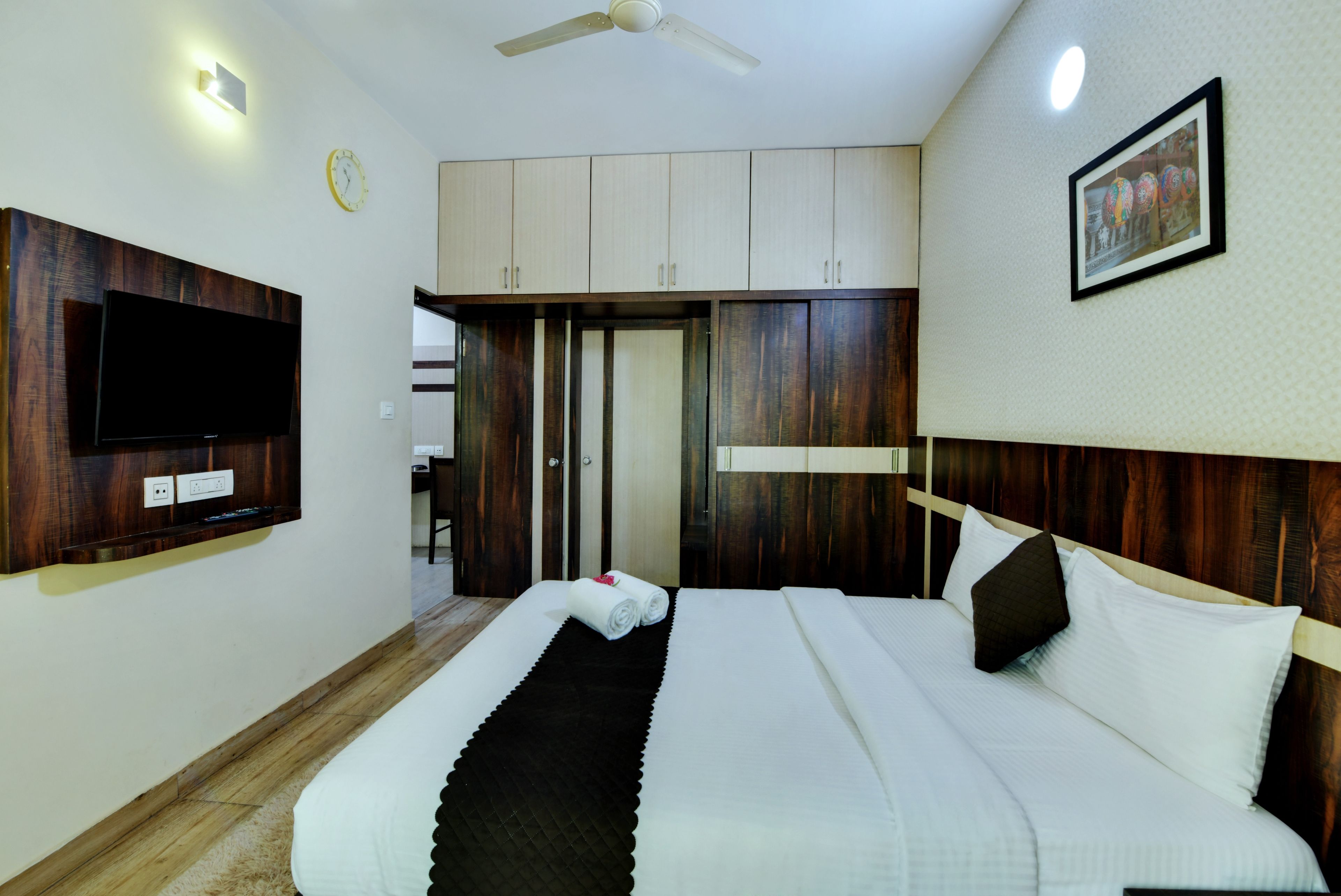 Quarto de hotel com uma cama, televisão de tela plana, armários de madeira, ventilador de teto e um quadro pendurado na parede.