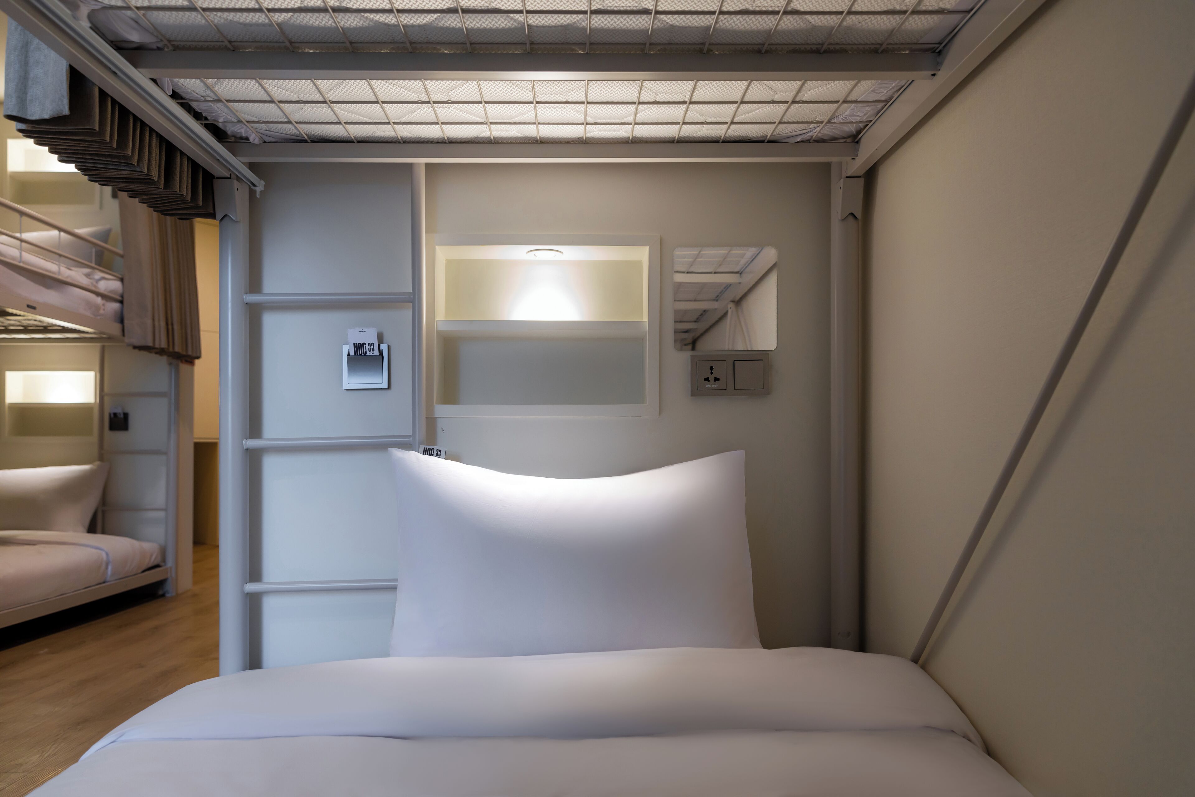 Quarto de hotel com uma cama beliche, uma mesinha de cabeceira, uma luminária na parede e um espelho.