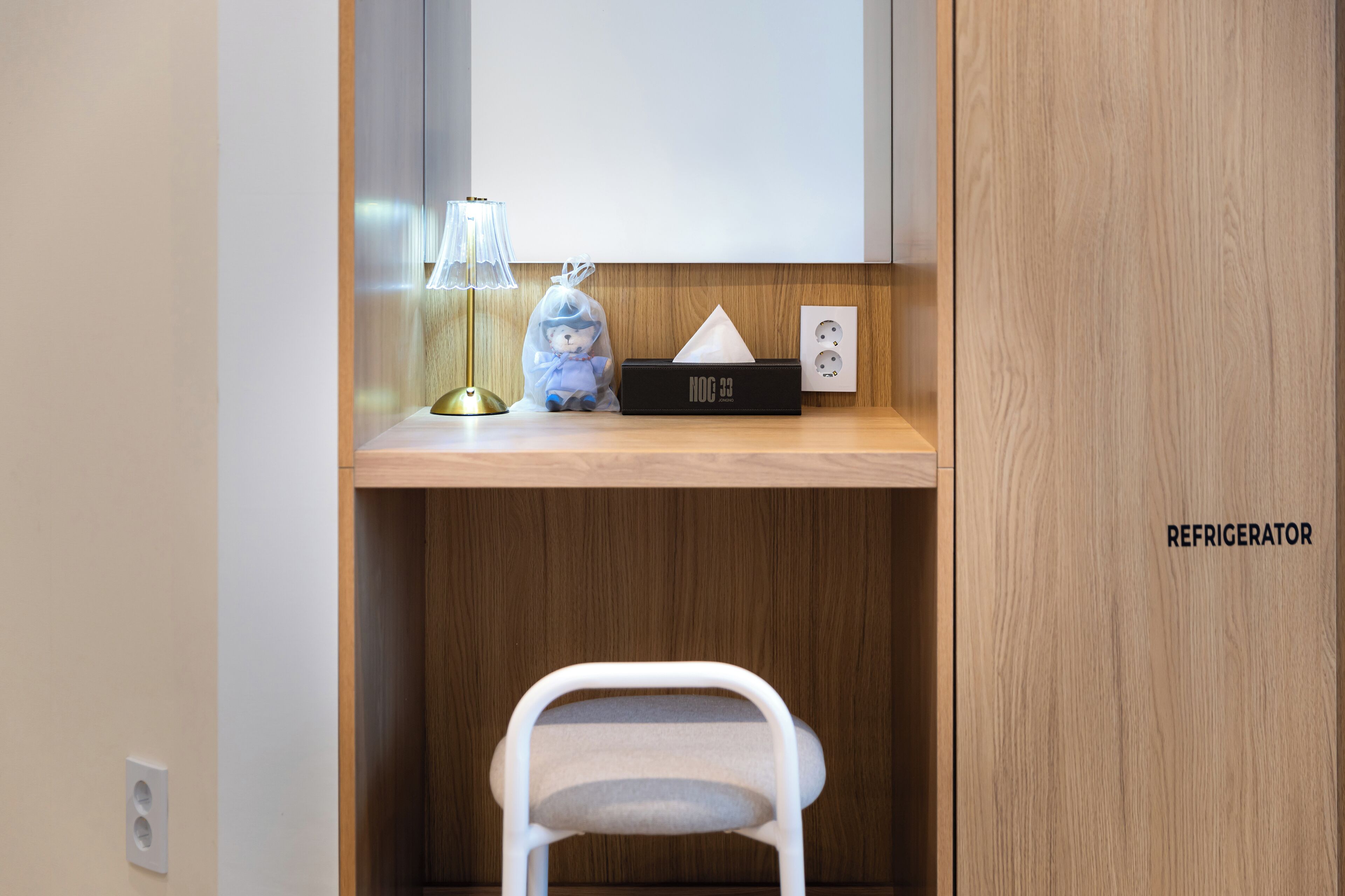 Uma área de cozinha compacta com bancada de madeira, uma cadeira pequena, uma luminária, um urso de pelúcia e uma caixa com a etiqueta 'NOC 33'.