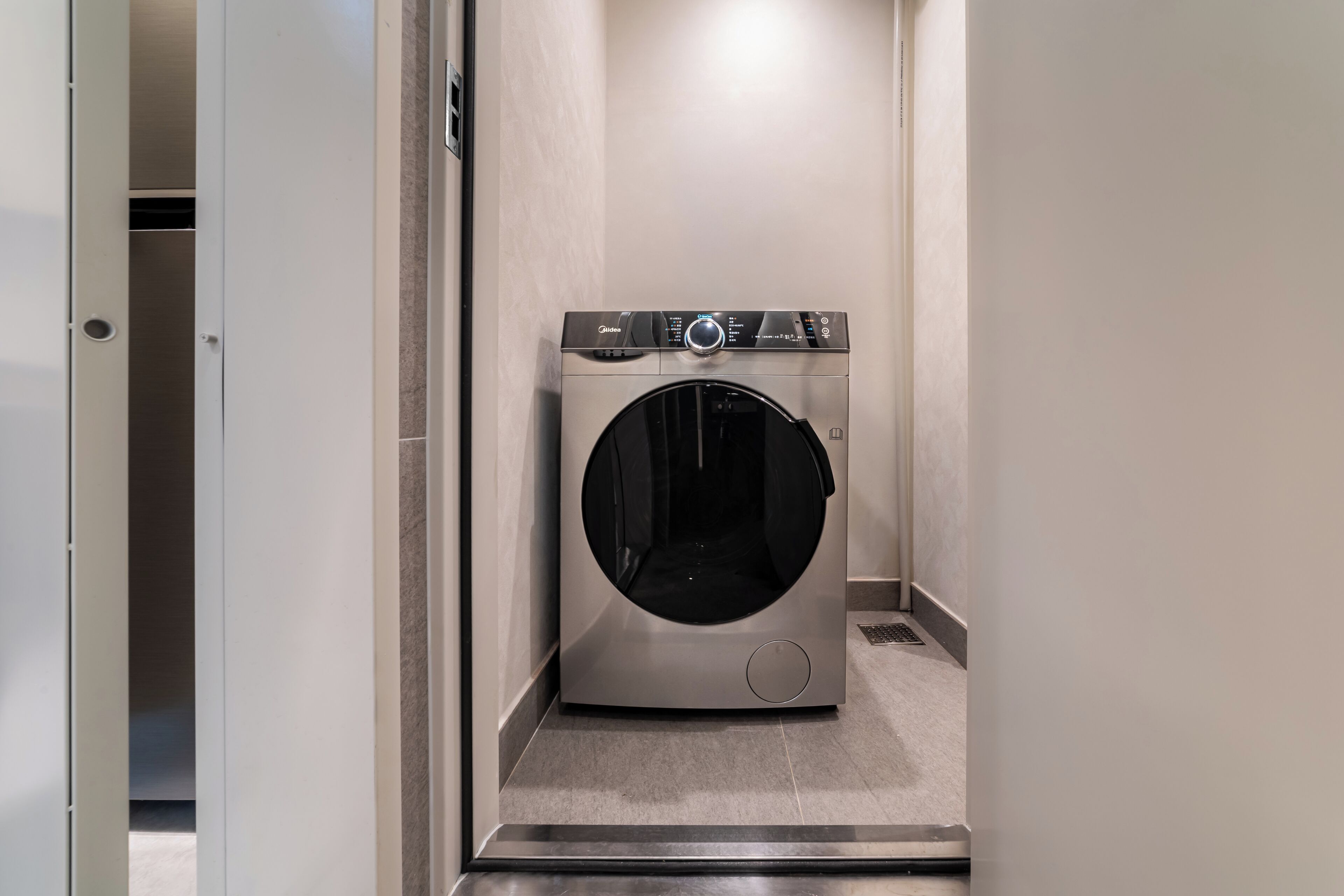 Máquina de lavar roupa de carregamento frontal, compacta e em aço inoxidável, numa pequena lavanderia fechada com chão em azulejo.