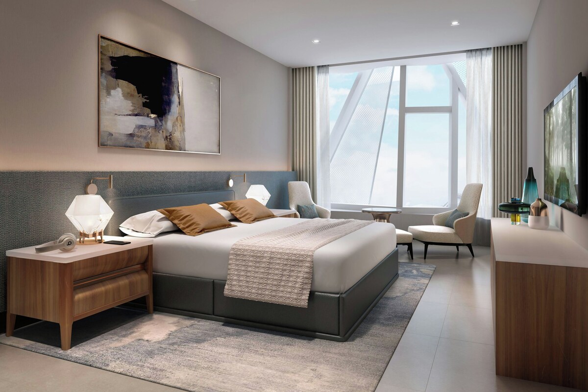 Quarto de hotel moderno com uma cama grande, uma escrivaninha, uma cadeira e uma janela ampla com cortinas.