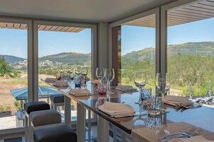 Uma área de jantar com mesa posta para refeição, grandes janelas com vista para o campo.