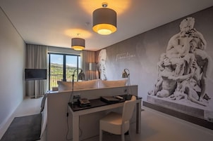 Quarto de hotel moderno com escrivaninha, uma janela grande, televisão de tela plana e uma escultura na parede.