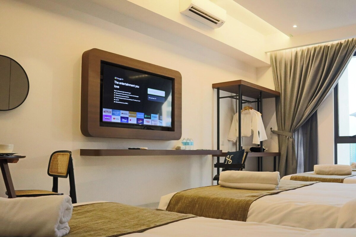 Quarto de hotel com duas camas, televisão fixada na parede, uma mesinha com cadeira e janela com cortinas.