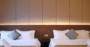 Duas camas com roupa de cama branca e almofadas cinzentas, uma cabeceira de madeira e um aplique na parede.