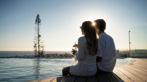 Um casal sentado à beira da piscina, com vista clara para o oceano e uma árvore ao longe.