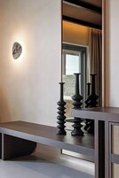 Um interior moderno com um aparador de madeira escura, um candeeiro fixo na parede e um espelho refletindo uma parede de pedra e uma janela com cortinas.