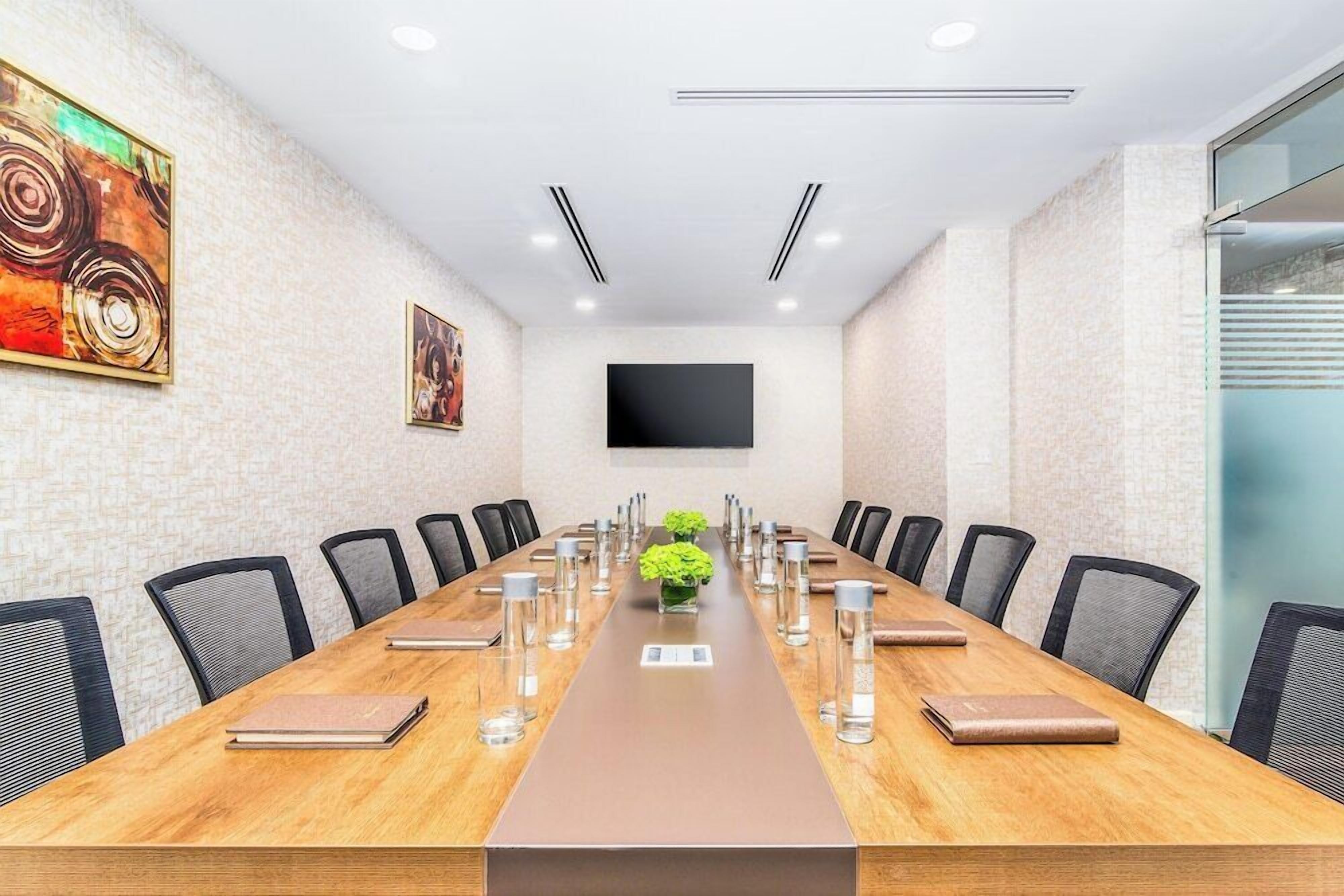 Uma sala de conferências com uma mesa comprida, cadeiras, uma TV de tela plana e um quadro na parede.