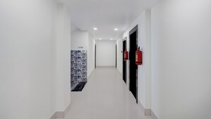 Um corredor comprido e estreito, com paredes brancas e chão de azulejos. Há portas de um lado e um extintor de incêndio fixado na parede.