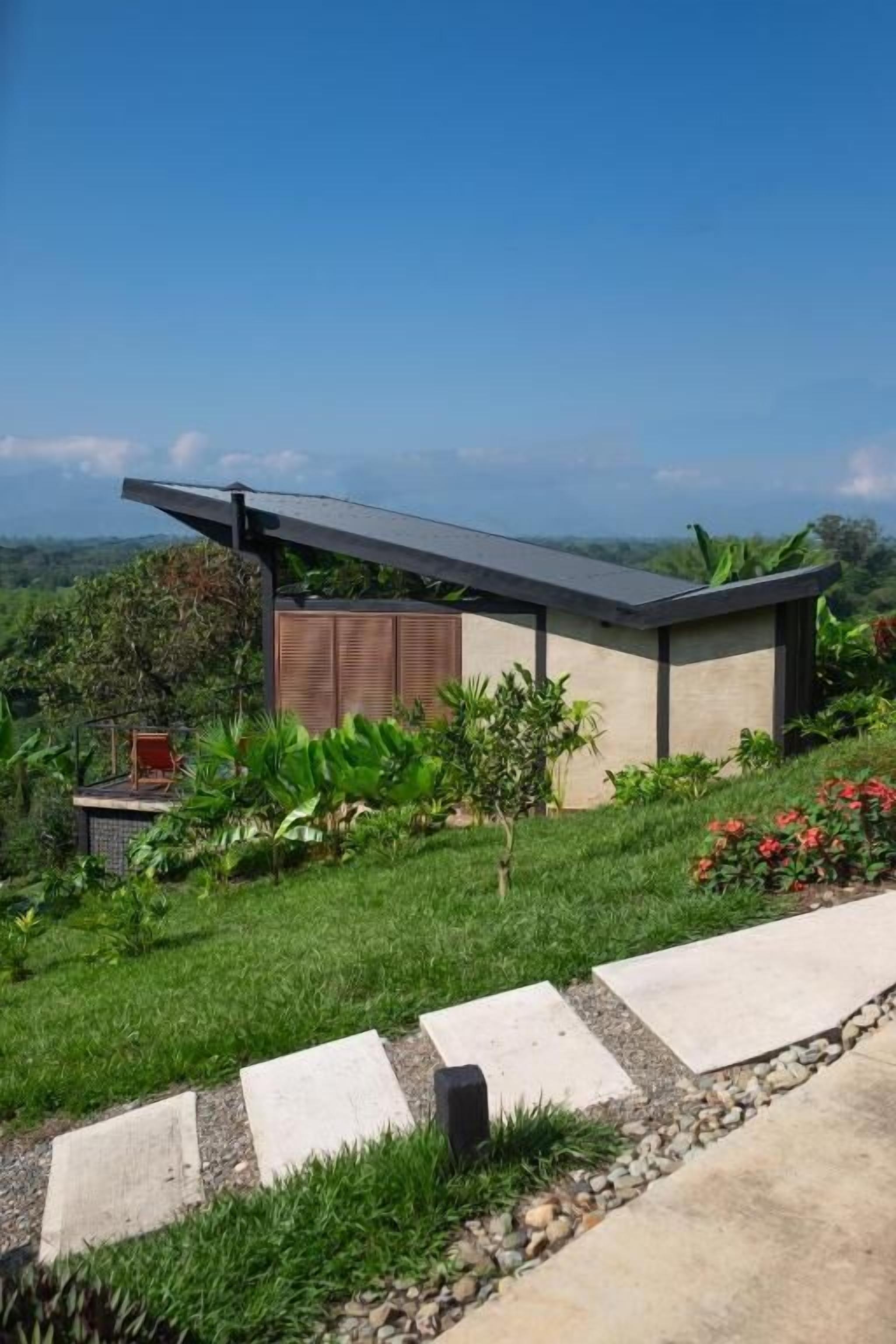 Um edifício moderno com telhado inclinado, rodeado por um jardim bem cuidado com diversas plantas e um caminho de pedra que leva à entrada.